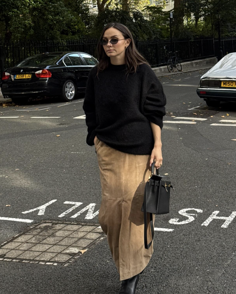 Woman wearing a black oversized sweater with a beige suede midi skirt%2C black ankle boots%2C small black leather handbag%2C and oval sunglasses in an urban setting